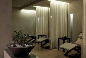 Um espaço de spa com uma grande bacia de pedra contendo uma árvore bonsai, duas poltronas de descanso, uma pequena mesa com um bule de chá e uma janela coberta por cortinas.