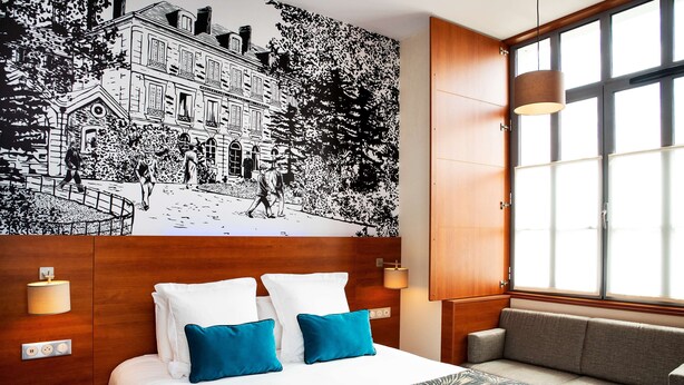 Quarto de hotel com uma cama grande, duas almofadas azuis, cabeceira de madeira, um mural na parede, um sofá e uma janela com vista.