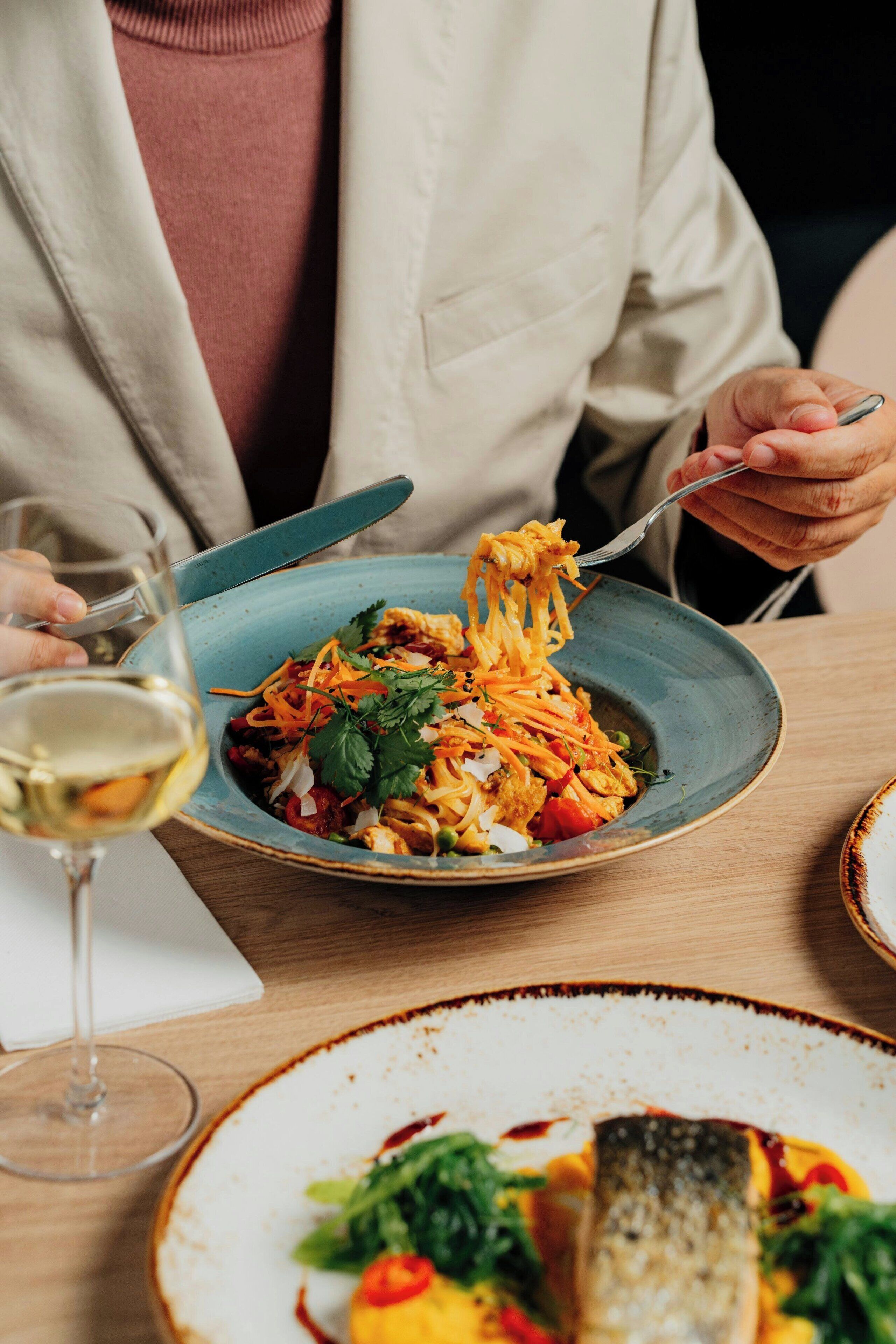Uma pessoa jantando à mesa, com um prato de comida e um copo de vinho branco.