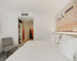 Quarto de hotel com cama, uma cadeira, um guarda-roupa e um mural de um edifício.