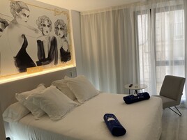 Quarto de hotel com uma cama grande, almofadas brancas, uma mesinha redonda com um controle remoto, uma cadeira e um quadro na parede.