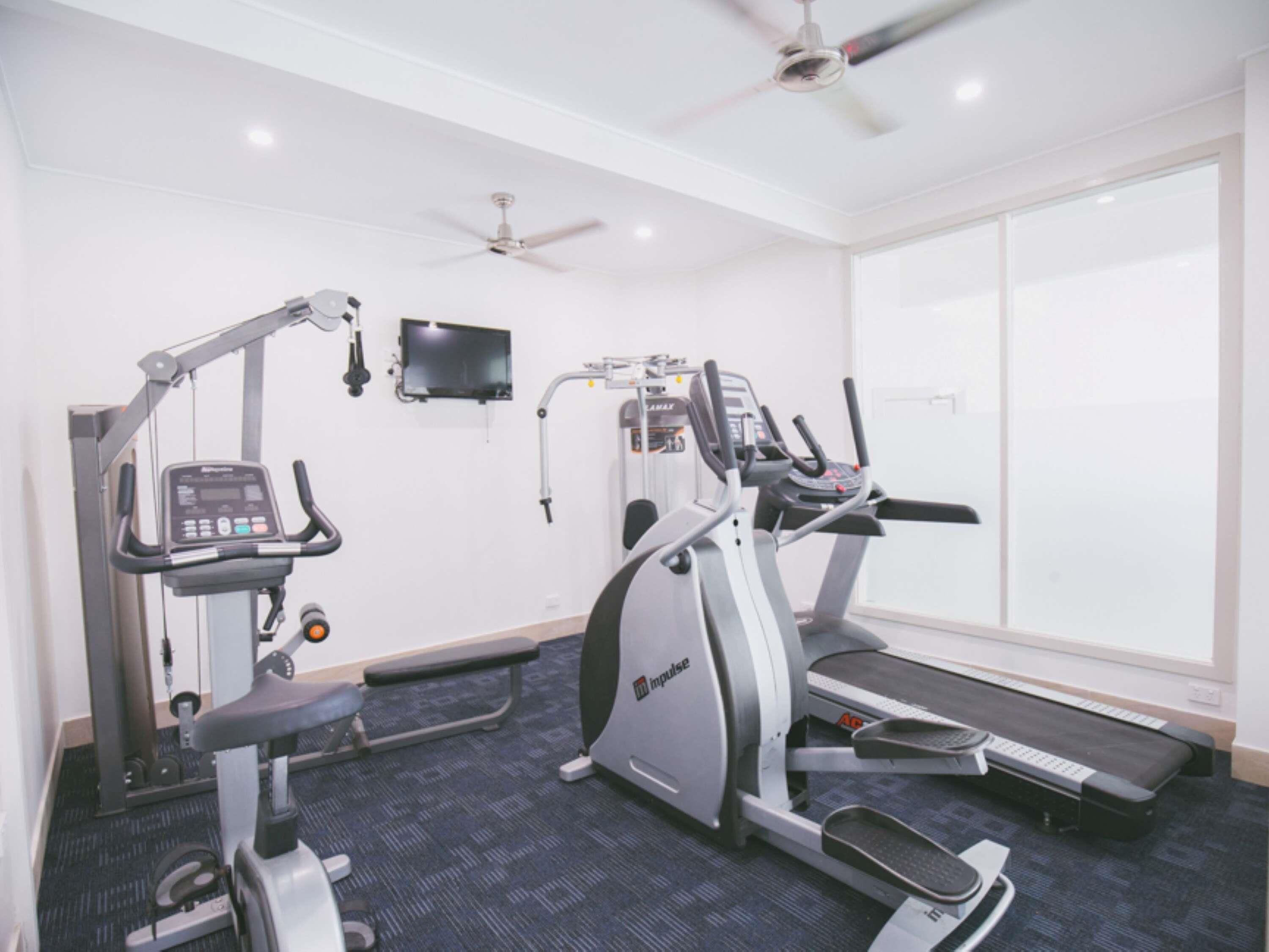 Academia com aparelhos de cardio, televisão e ventilador de teto.