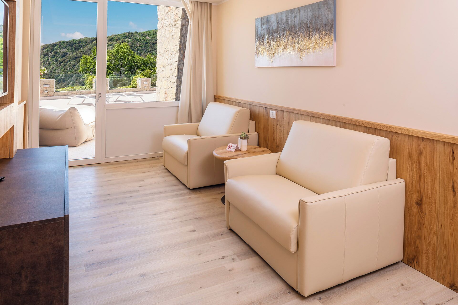 Um quarto com dois sofás bege, piso de madeira, um quadro na parede e vista para o verde lá fora.