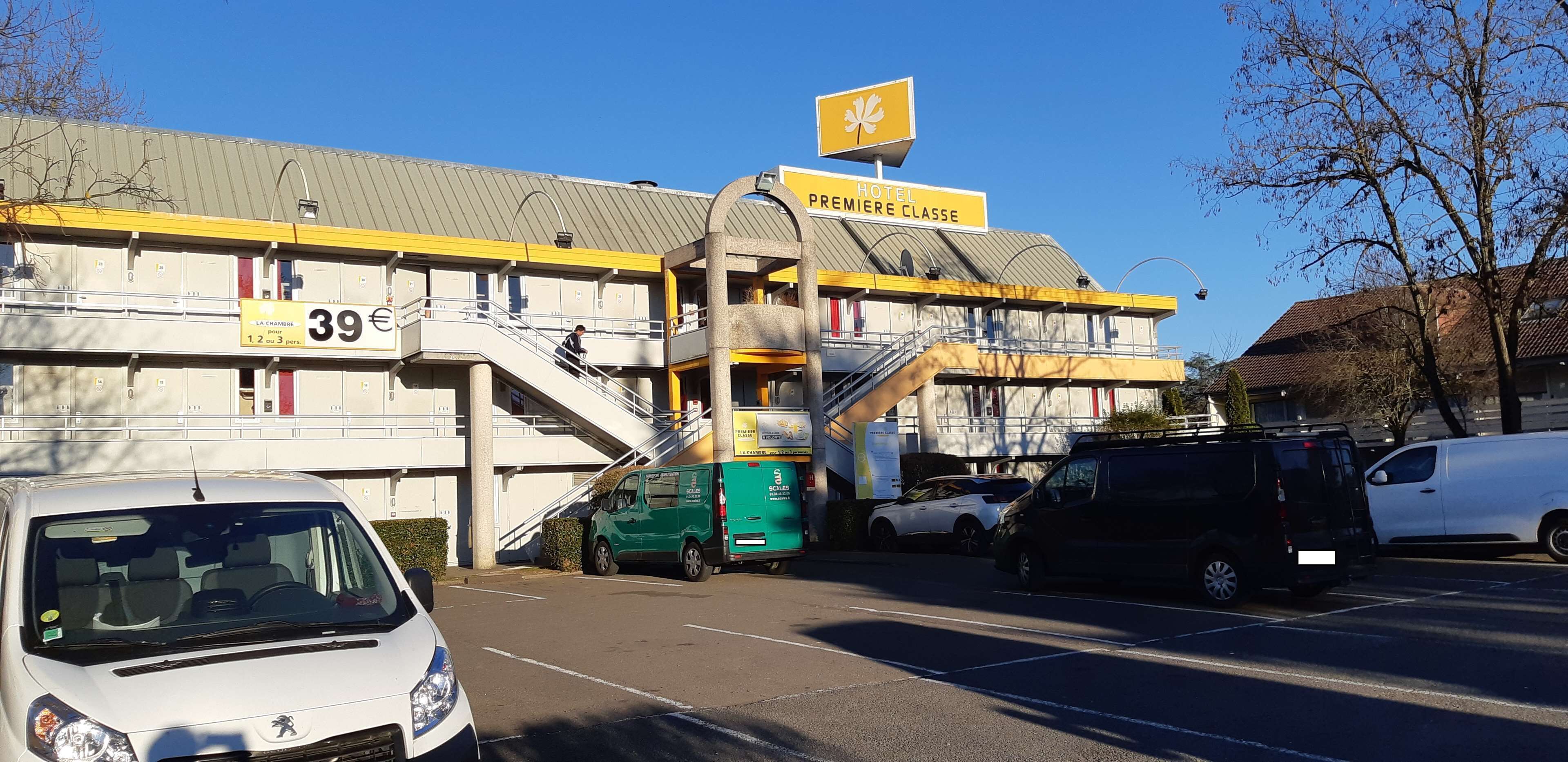 Hotel com placa indicando "HOTEL PREMIERE CLASSE" e etiqueta de preço com a inscrição "39€". Vários veículos estão estacionados na frente, incluindo um carro branco e uma van verde.