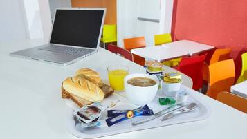 Bandeja de café da manhã com pão, suco, café, iogurte e acompanhamentos sobre uma mesa branca em área de refeições colorida, com cadeiras laranja e um laptop sobre a mesa.