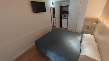 Quarto compacto com uma cama, televisão na parede, cozinha compacta e uma janela pequena.