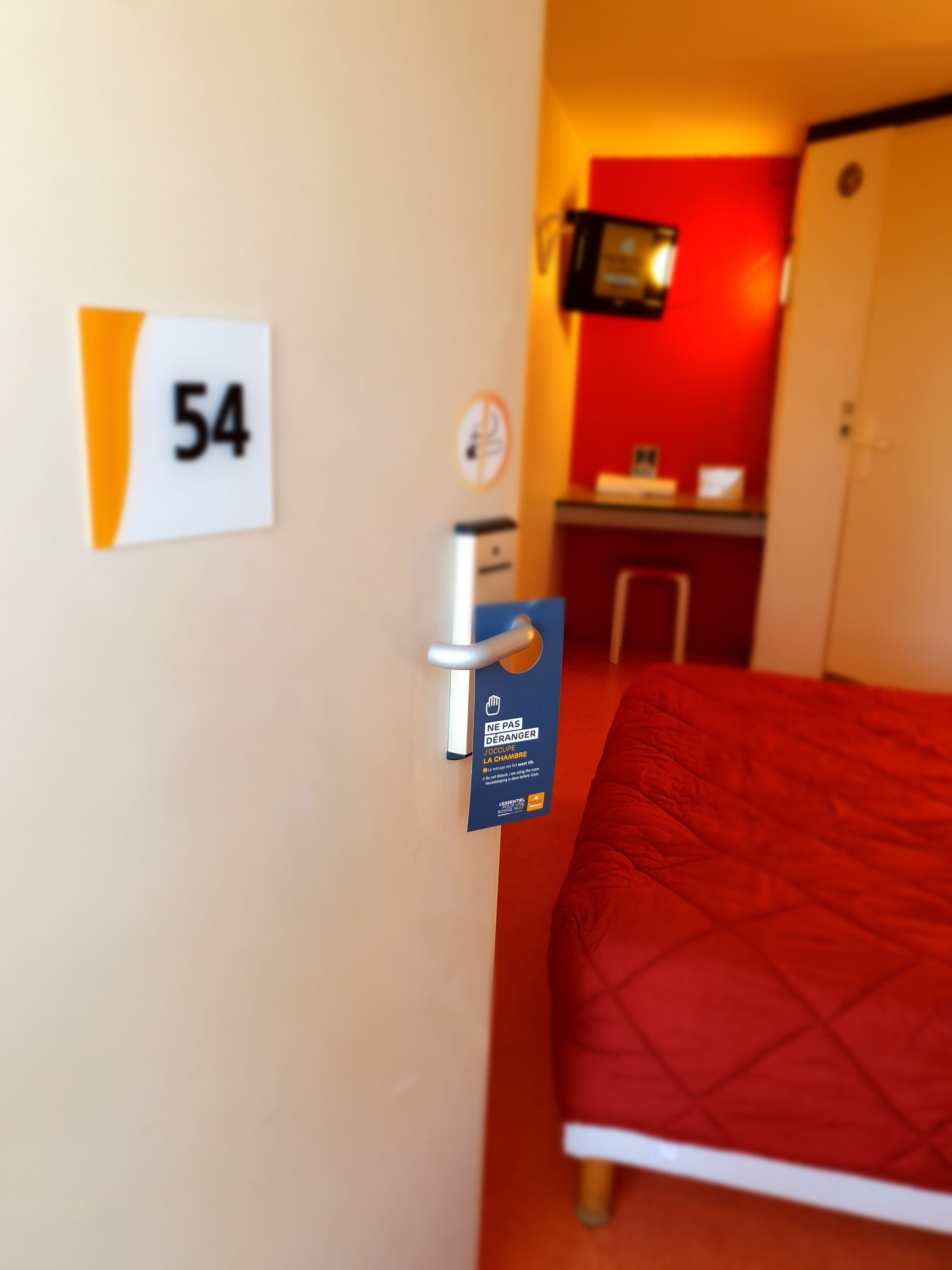 Quarto de hotel com cama, porta com placa de número, frigobar e televisão na parede.