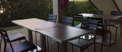 Uma área de refeições ao ar livre com mesas e cadeiras, uma estrutura de tenda e um sebe ao fundo.