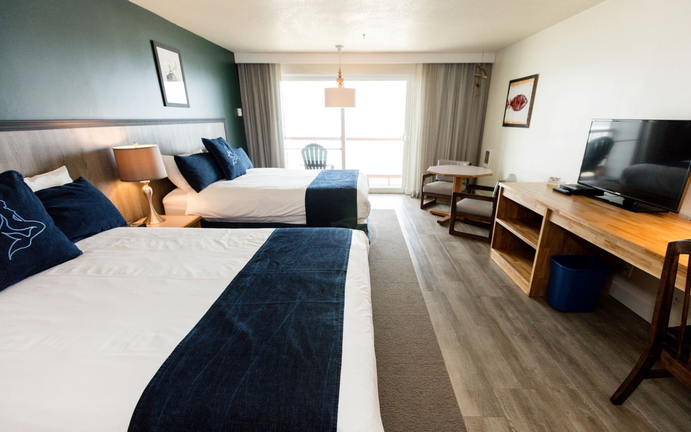 Quarto de hotel com duas camas, uma escrivaninha com televisão, uma cadeira e vista para o exterior.