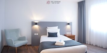 Quarto de hotel moderno com uma cama grande, uma poltrona azul clara e uma mesinha de cabeceira de madeira.