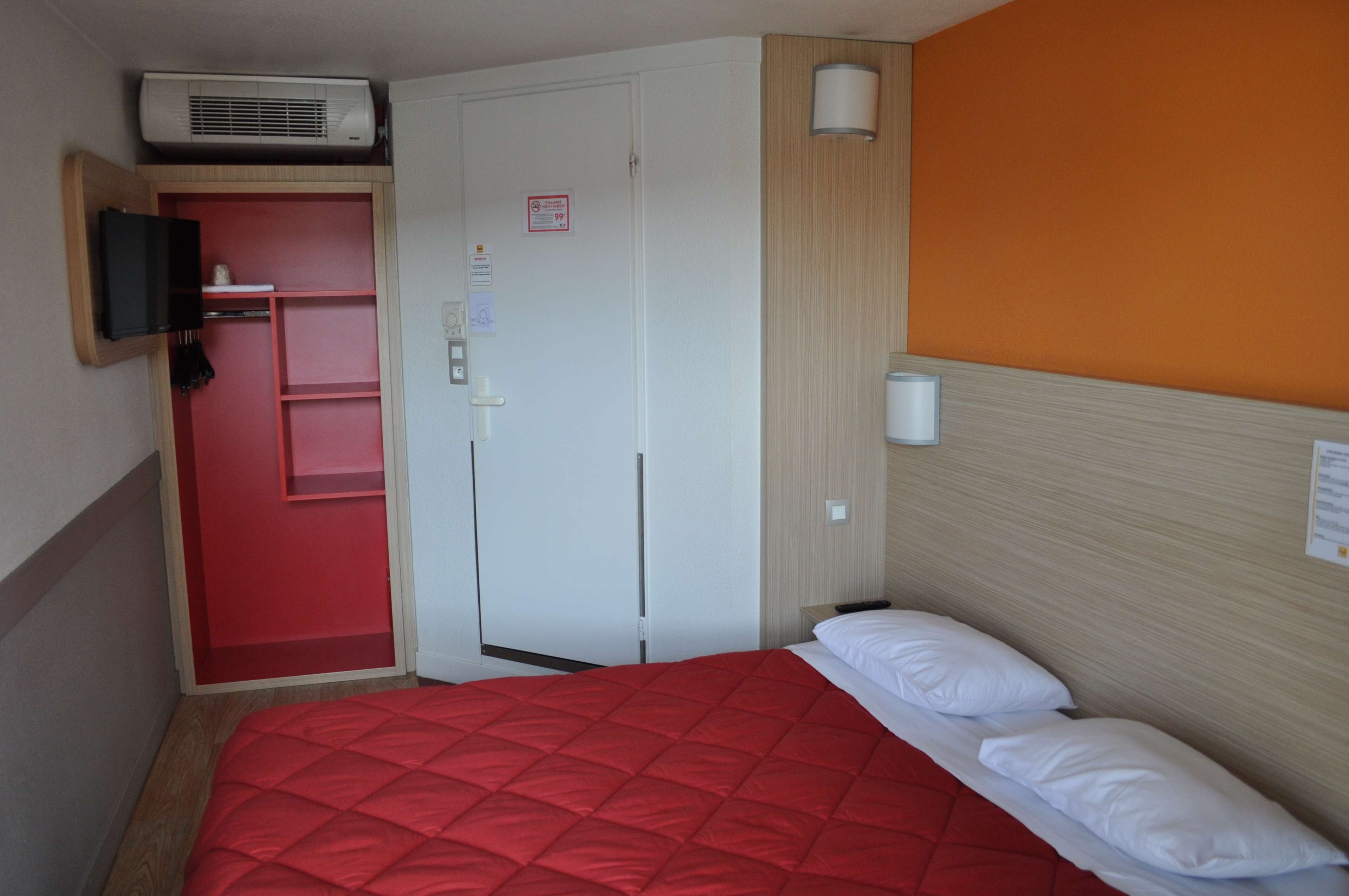 Quarto de hotel com colcha vermelha, travesseiro branco, guarda-roupa vermelho e televisão fixada na parede.