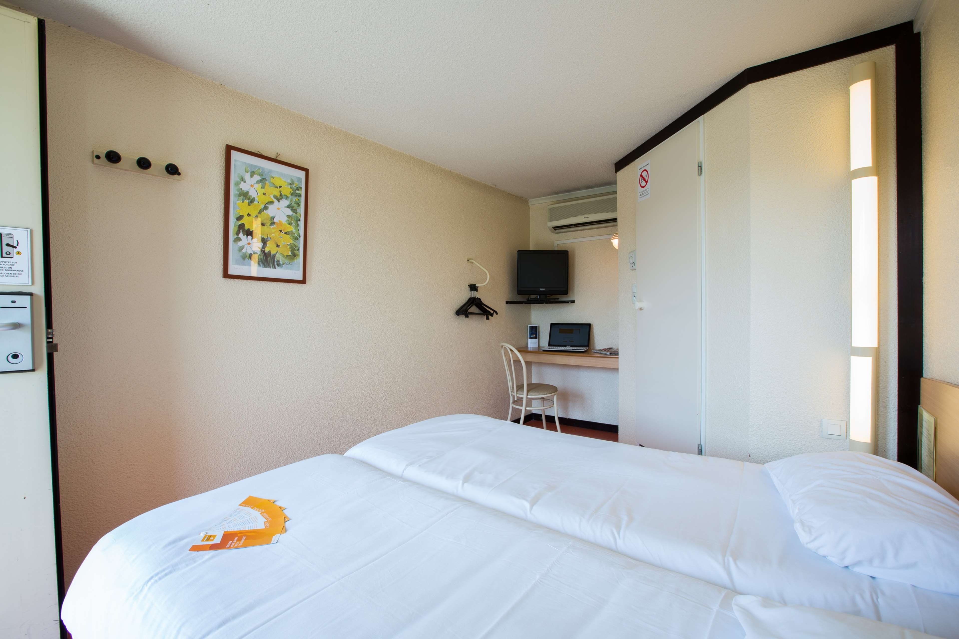 Quarto de hotel com cama, escrivaninha com computador, televisão e ar condicionado fixado na parede.