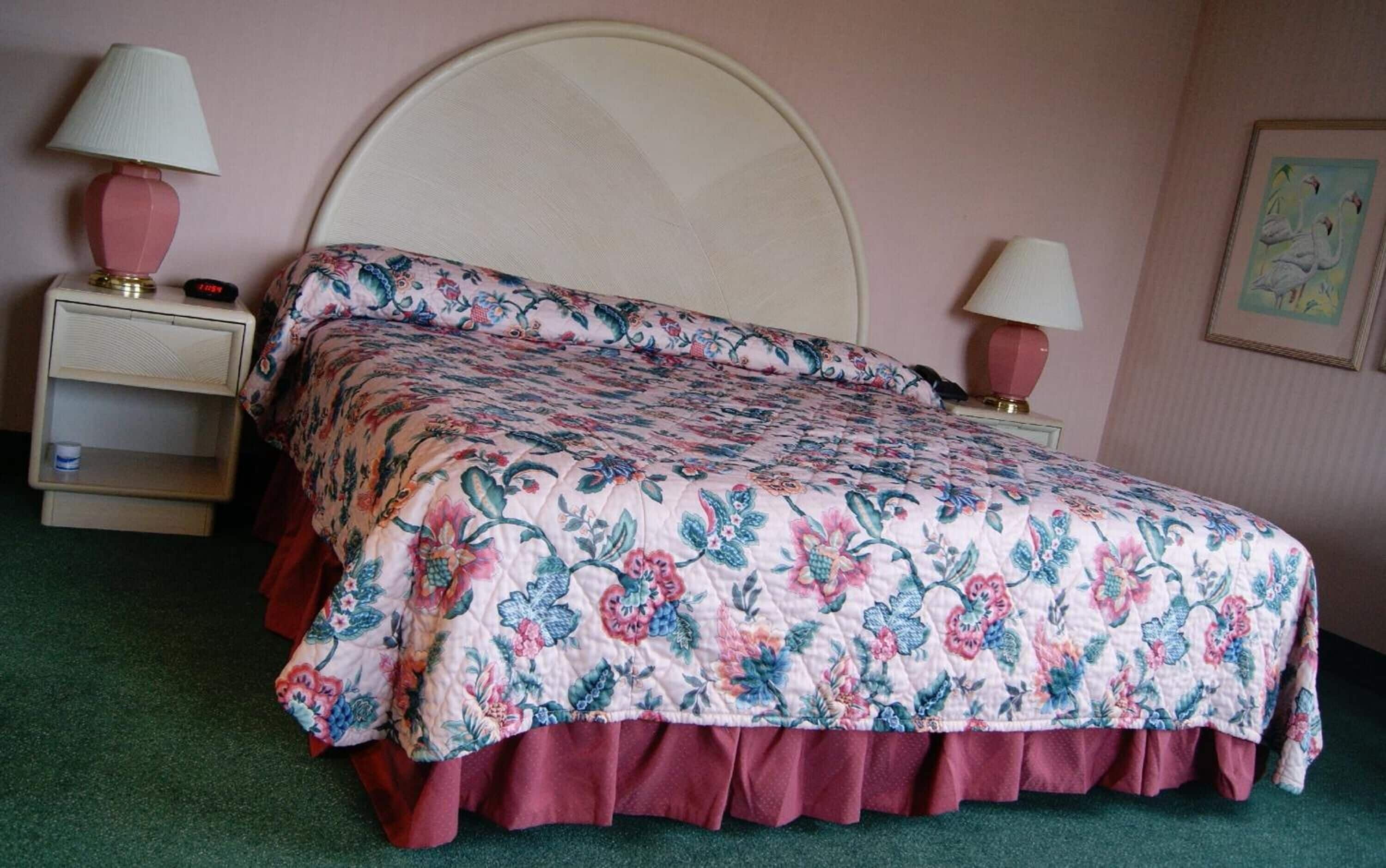 Uma cama com colcha florida, duas luminárias de cabeceira, mesa de cabeceira com relógio e um quadro na parede.