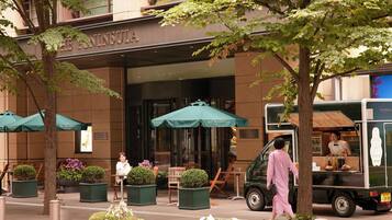 Entrada de hotel com área de estar externa, food truck e plantas em vasos.