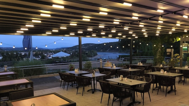 Um restaurante ao ar livre com mesas e cadeiras, vista para um rio e colinas ao fundo, e luzes penduradas por cima.