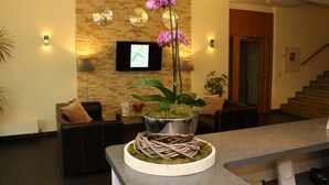 Uma área de recepção com balcão, uma orquídea em vaso, parede de pedra com TV fixada e escada ao fundo.