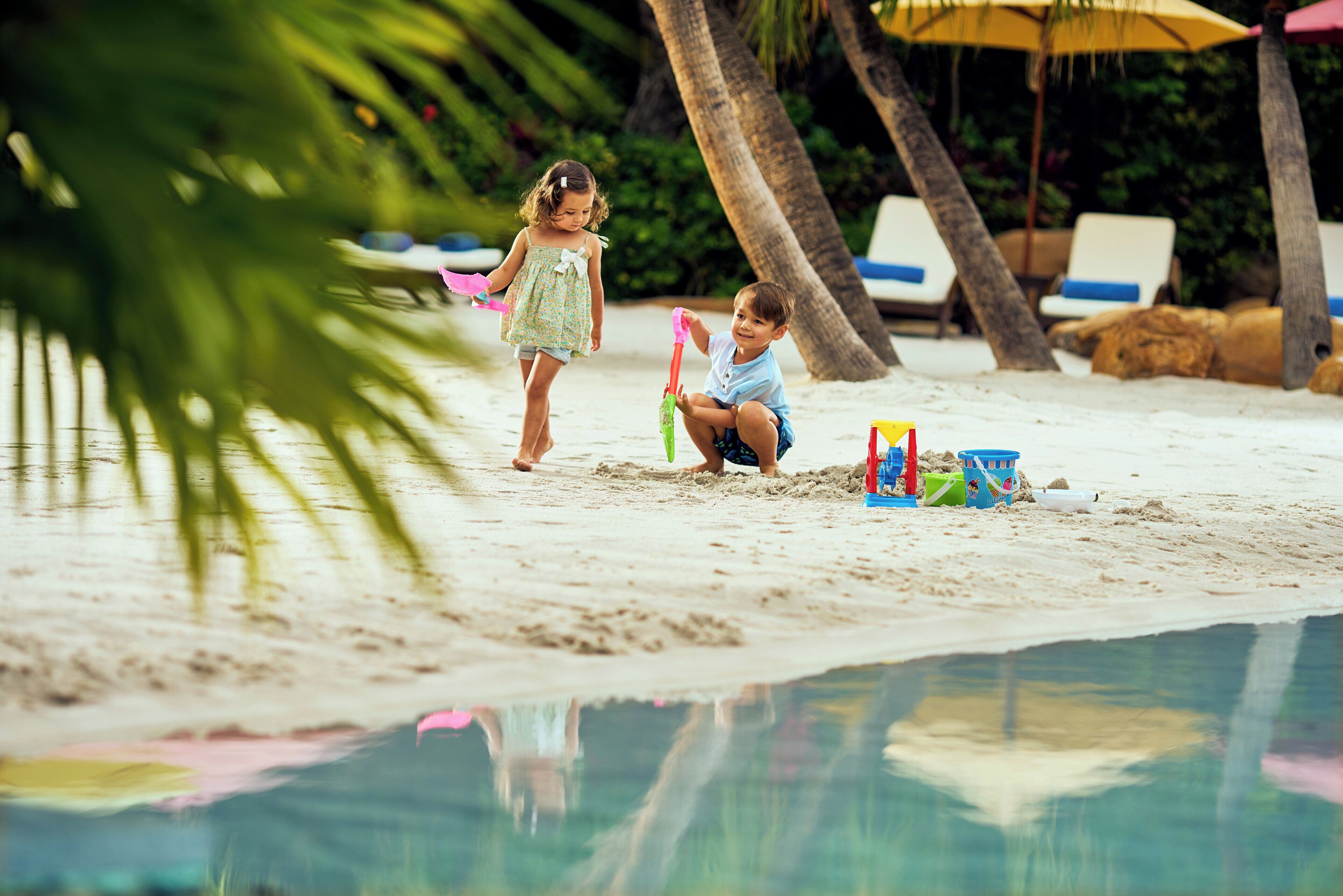 Duas crianças brincam em uma praia arenosa, com uma piscina ao fundo.