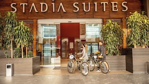 A entrada do Stadia Suites com um design moderno, apresentando fachada de madeira e grandes vasos de plantas.