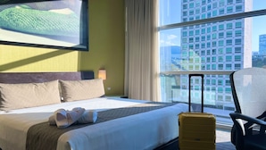 Quarto de hotel com uma cama, uma mala, uma cadeira e uma janela grande com vista para a cidade.