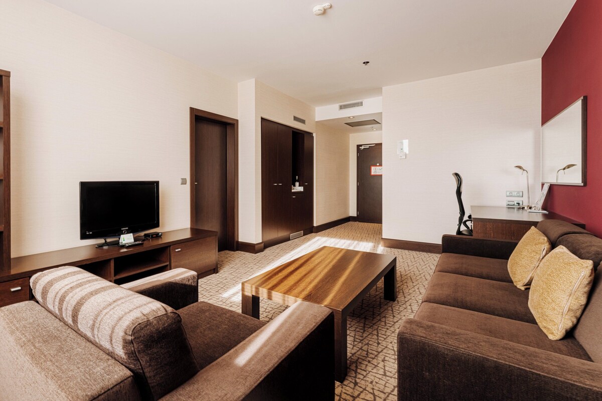 Quarto de hotel moderno com televisão de tela plana, móvel de entretenimento em madeira, sofá, mesa de centro e parede com destaque em vermelho.