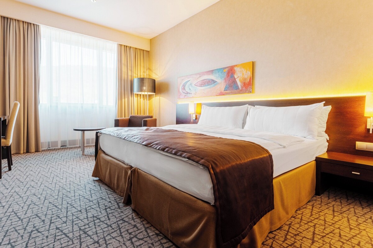 Quarto de hotel com uma cama, escrivaninha, cadeira, luminária e um quadro na parede.