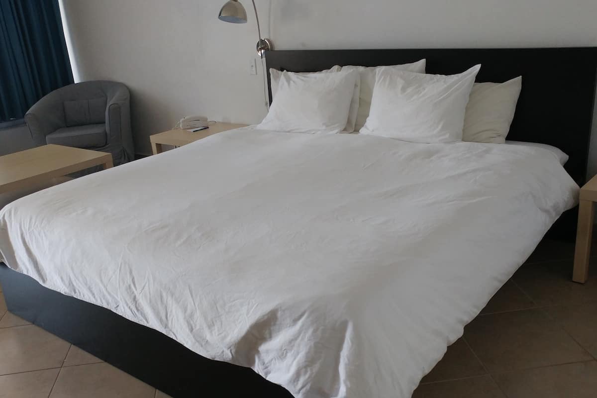 Uma cama grande com roupa de cama branca, uma mesa de cabeceira, uma luminária e uma cadeira em um quarto de hotel.