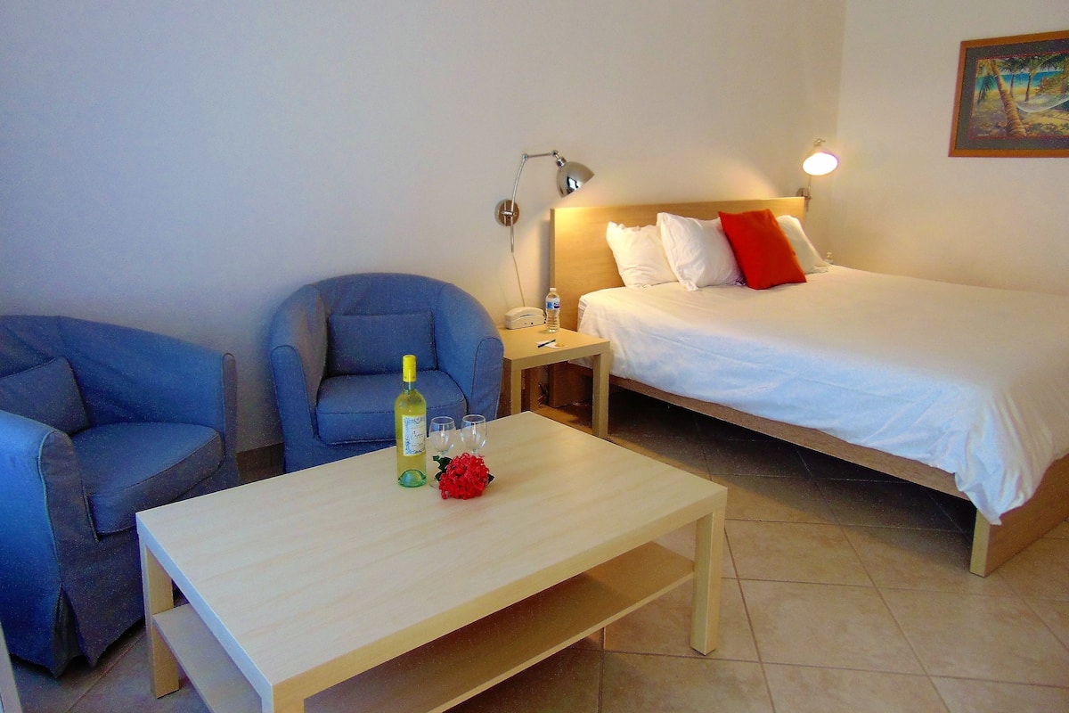 Quarto de hotel com cama, poltrona azul, mesa de madeira com uma garrafa e copos, e mesa de cabeceira com abajur.
