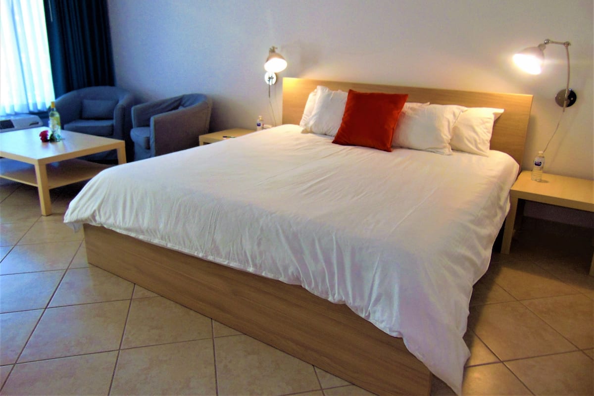 Quarto de hotel com uma cama grande, duas mesas de cabeceira, um criado-mudo, um sofá e uma mesa de centro.