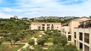 Um edifício de vários andares com varandas, rodeado por oliveiras e uma paisagem montanhosa ao fundo.