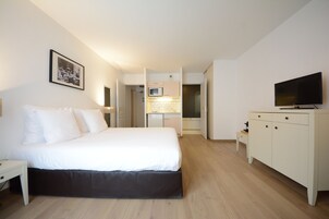 Quarto de hotel com cama, televisão, escrivaninha e cozinha compacta.