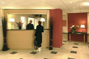 Uma área de recepção com um balcão de madeira, um espelho e duas luminárias. Há plantas decorativas em ambos os lados do balcão.