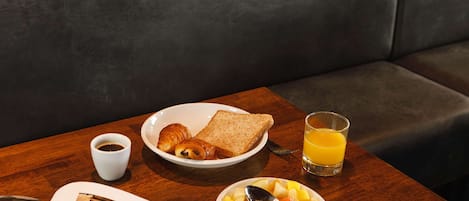 Disposição de café da manhã com croissants, torradas, suco de laranja e uma cesta de frutas sobre uma mesa de madeira.