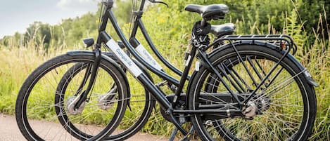 Uma bicicleta preta com cesto frontal e bagageiro traseiro, estacionada em um caminho pavimentado com vegetação ao fundo.