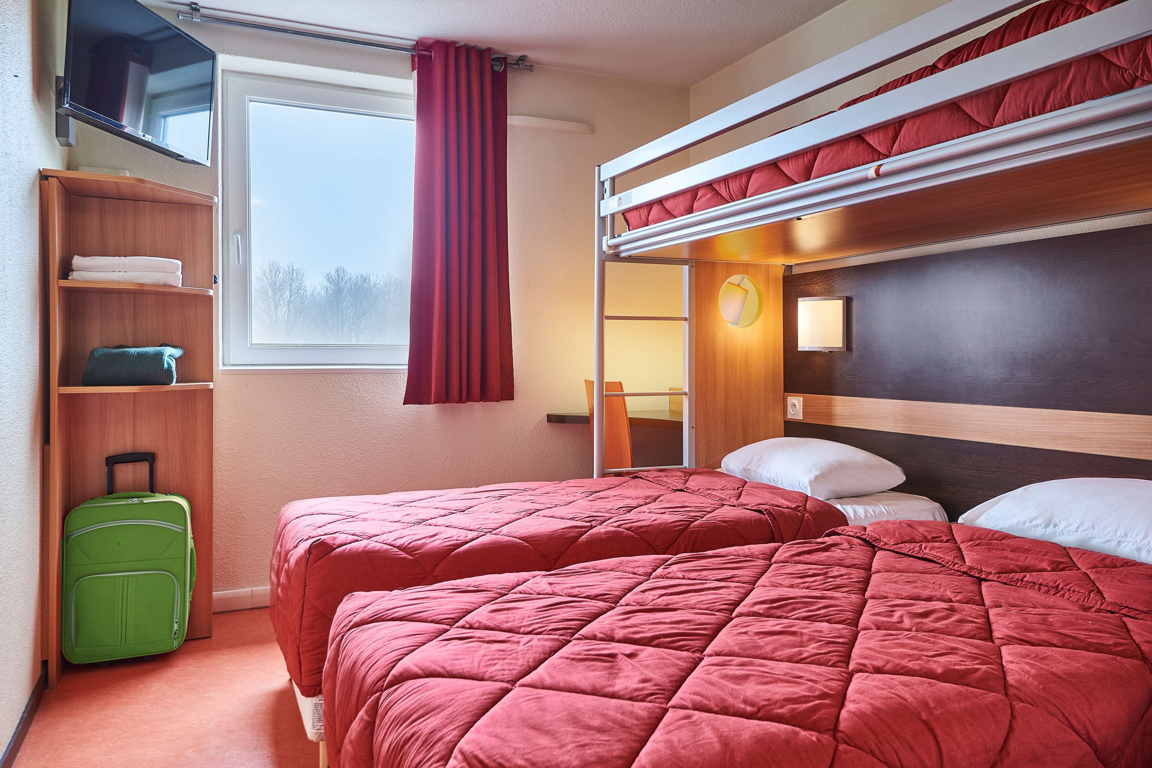 Quarto de hotel com beliches, uma mala verde, uma prateleira de madeira e uma janela com cortina vermelha.