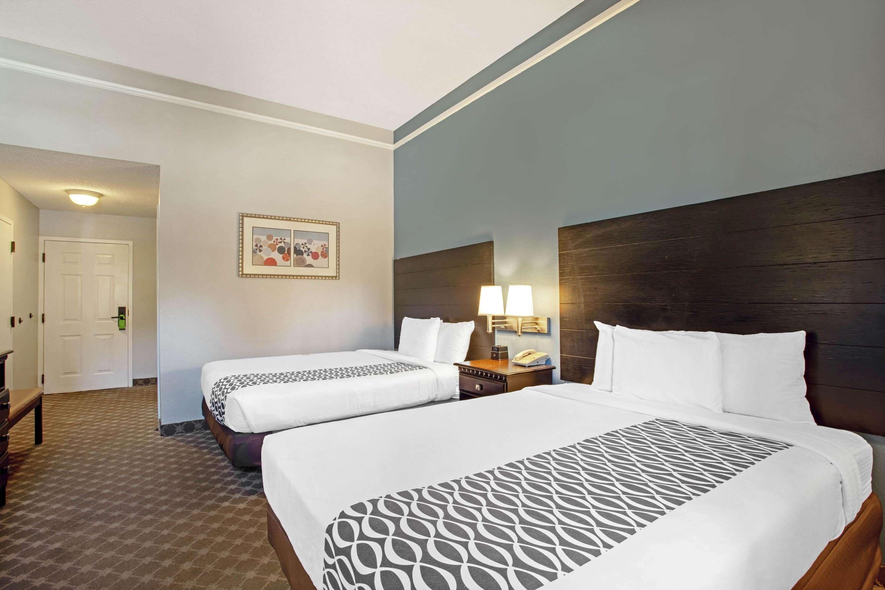 Quarto de hotel com duas camas, uma cabeceira de madeira, uma mesa de cabeceira com abajur e um quadro emoldurado na parede.
