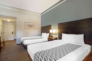 Quarto de hotel com duas camas, uma cabeceira de madeira, uma mesa de cabeceira com abajur e um quadro emoldurado na parede.