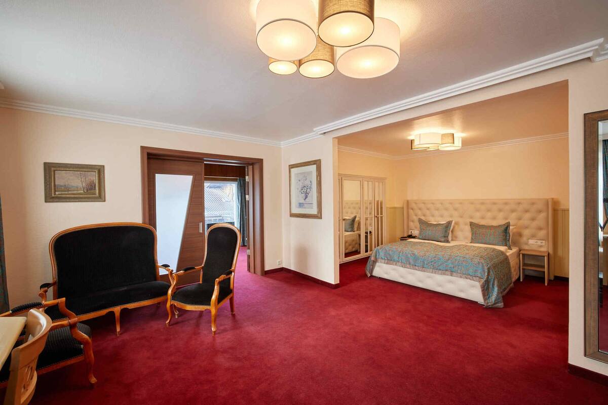Quarto de hotel com tapete vermelho, uma cama grande com colcha estampada, duas poltronas estofadas, uma mesa lateral de madeira, um criado-mudo e uma janela ampla com vista.