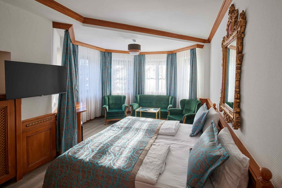 Quarto de hotel com uma cama grande, duas poltronas verdes, uma mesa pequena, uma TV e um espelho grande e ornamentado.
