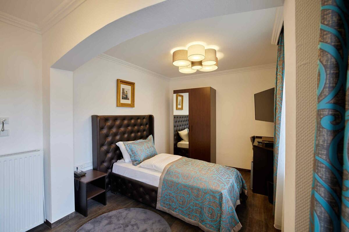 Quarto com cabeceira estofada, cama com roupa de cama estampada, mesa de cabeceira, guarda-roupa, espelho e luminária de teto.