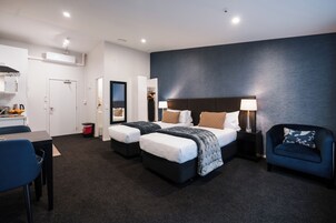 Quarto de hotel com duas camas, uma escrivaninha, uma cadeira, um guarda-roupa e uma mesa de cabeceira com abajur.
