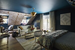 Quarto moderno com uma cama grande, um sofá e uma mesa de jantar. Há janelas com cortinas, um lustre e uma peça decorativa na parede.