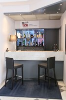 Uma área de bar com balcão de mármore, dois banquetas pretas, uma luminária e uma estante com vários frascos.