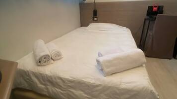 Quarto de hotel com cama, mesa de cabeceira, uma cadeira, televisão fixada na parede e um quadro emoldurado.