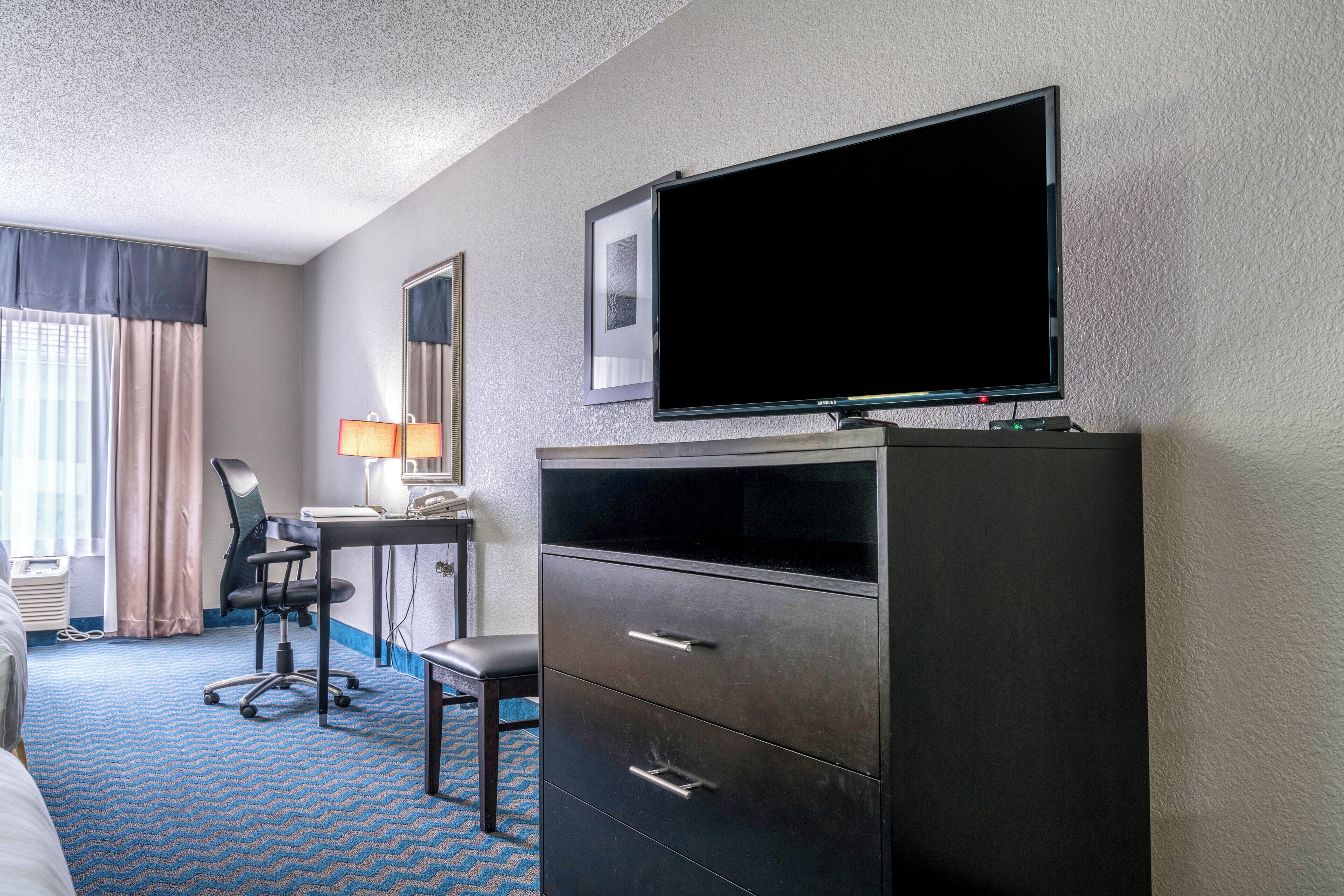 Quarto de hotel com cama, escrivaninha com cadeira, televisão fixada na parede e um armário grande.