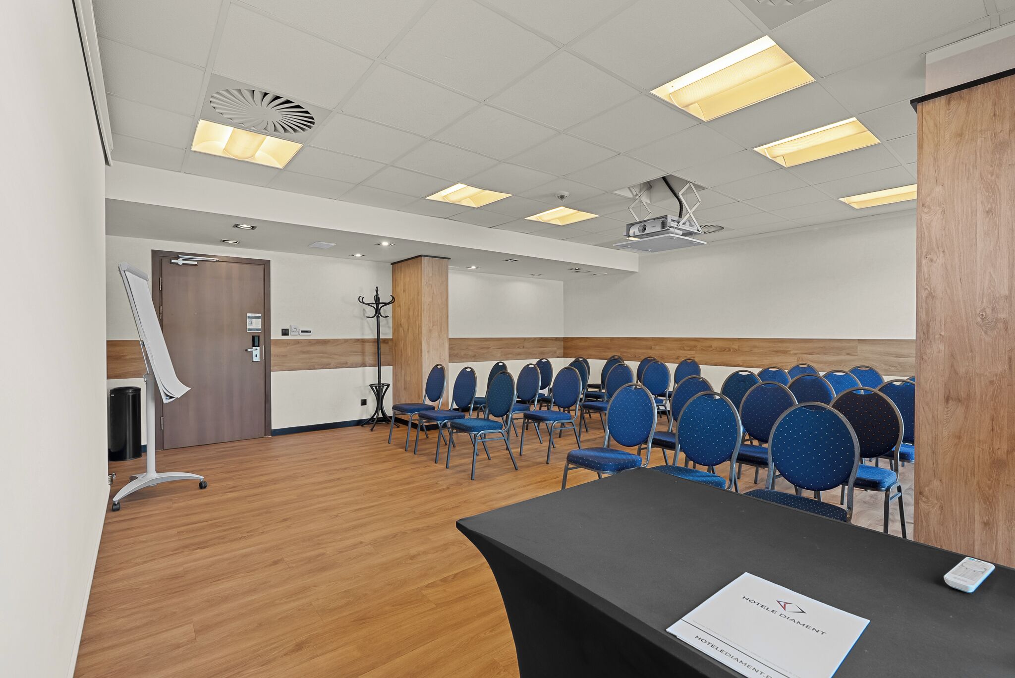 Sala de conferências com uma mesa comprida, fileiras de cadeiras, um projetor e um cabideiro.