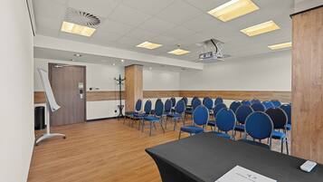 Sala de conferências com uma mesa comprida, fileiras de cadeiras, um projetor e um cabideiro.