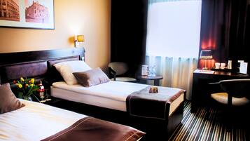 Quarto de hotel com duas camas, uma escrivaninha, uma cadeira, uma mesinha com garrafas de água e um abajur.