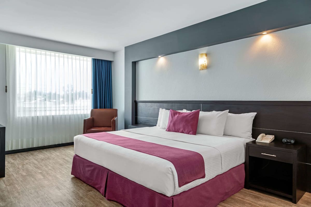 Quarto de hotel com uma cama grande, uma mesa de cabeceira, uma cadeira e uma janela com cortinas.