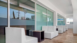Espaço interior moderno com sofás brancos e piso ladrilhado, grandes janelas e vista para o exterior.