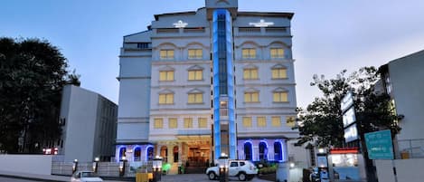 Um hotel com vários andares, com um eixo de elevador de vidro azul central, diversas varandas e uma entrada bem iluminada.